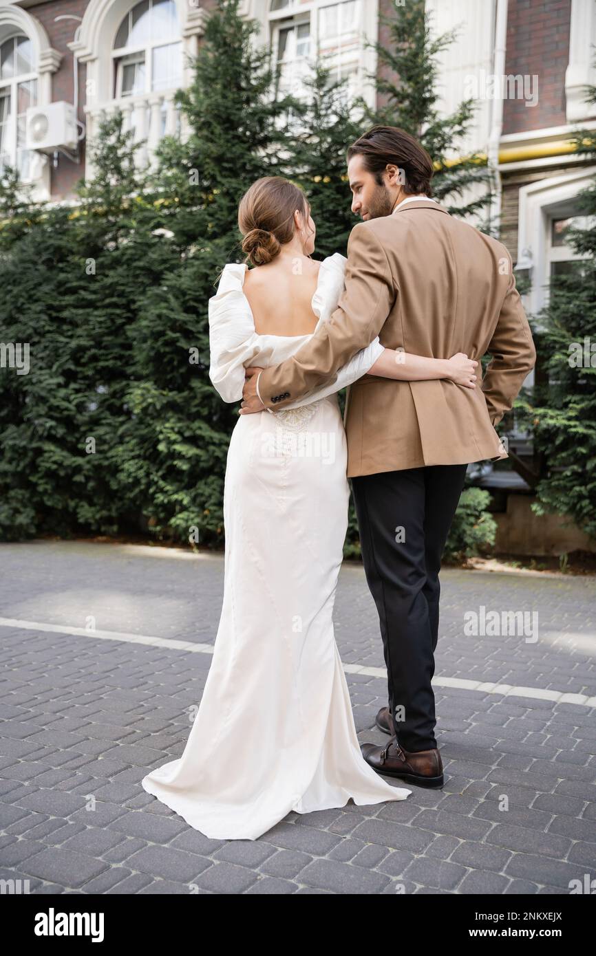 back view of bride in white dress hugging with bearded groom on street ...