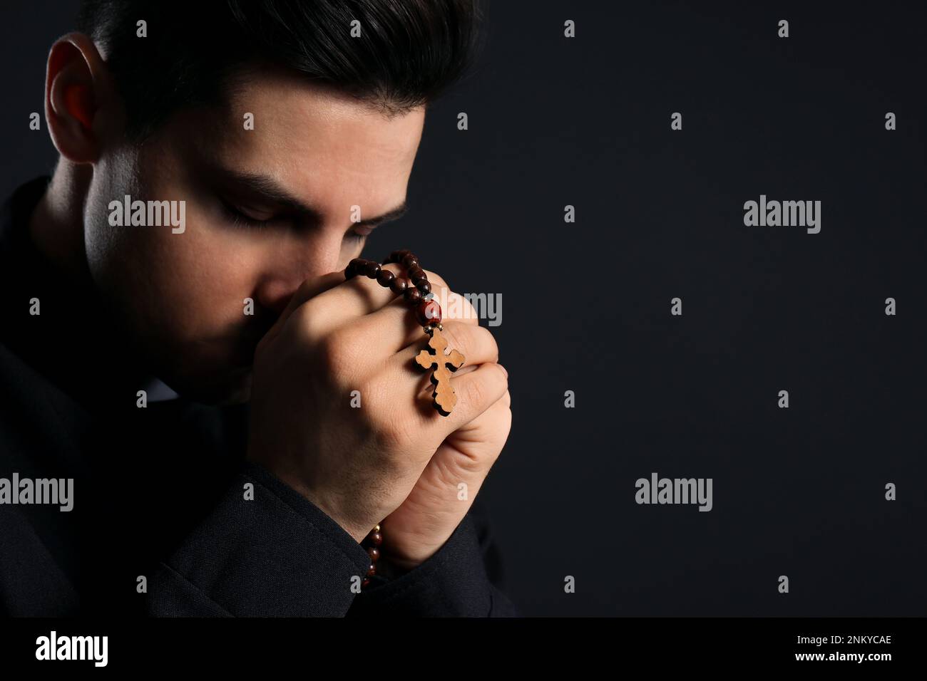 Priest with rosary beads praying on black background, closeup. Space ...
