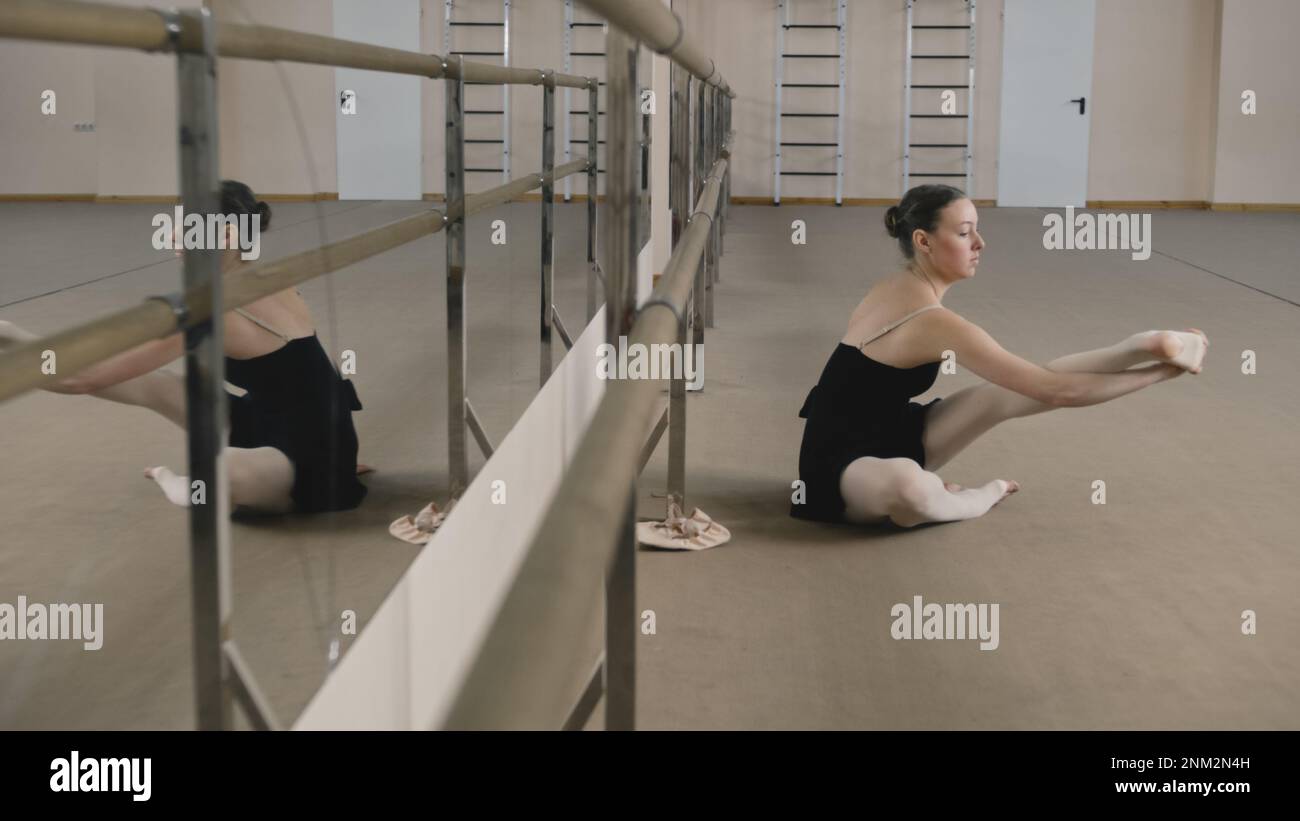 Ballet dancer in training suit near ballet barre stretches legs before ...