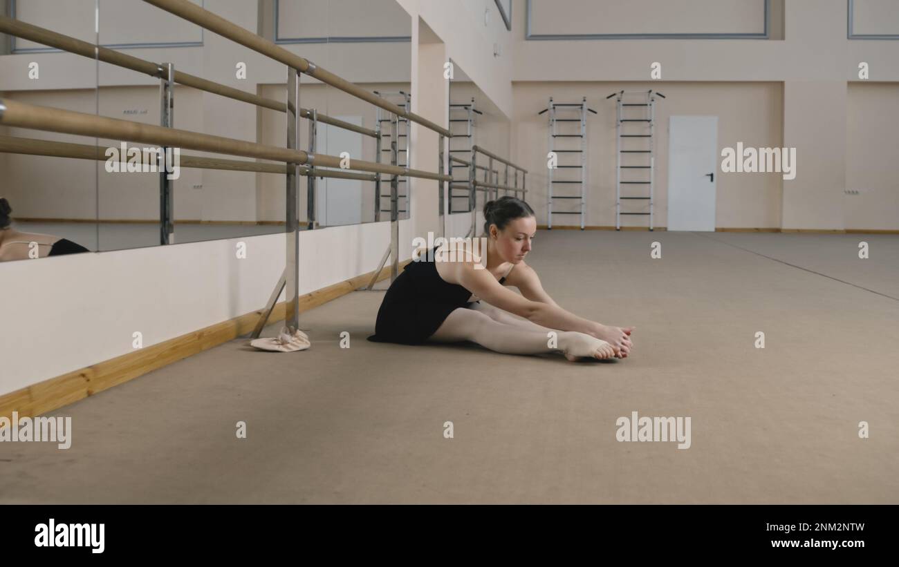Ballet dancer in training suit near ballet barre stretches legs before ...
