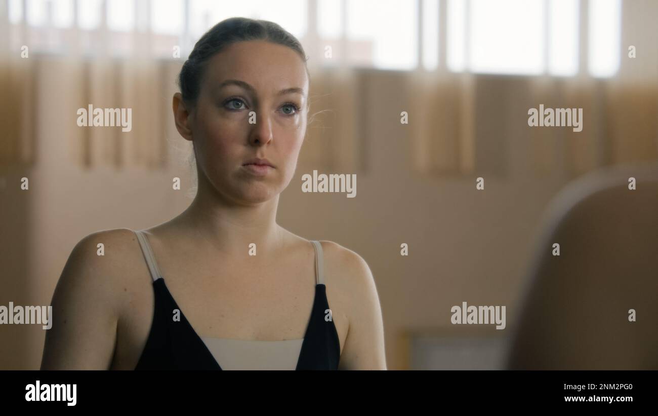 Tired ballerina in training bodysuit stands near ballet barre in dance ...