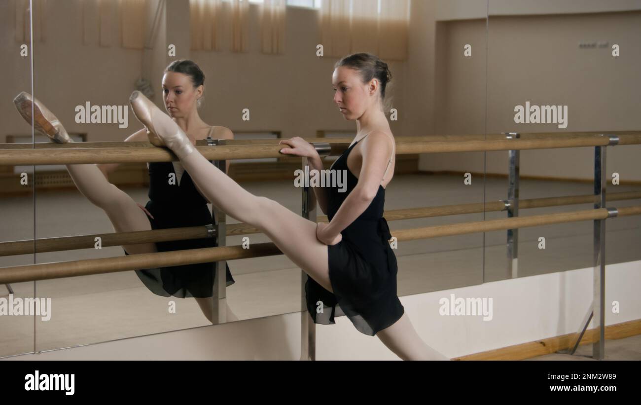 Tired ballerina in training bodysuit stands near ballet barre in dance studio during preparing ...