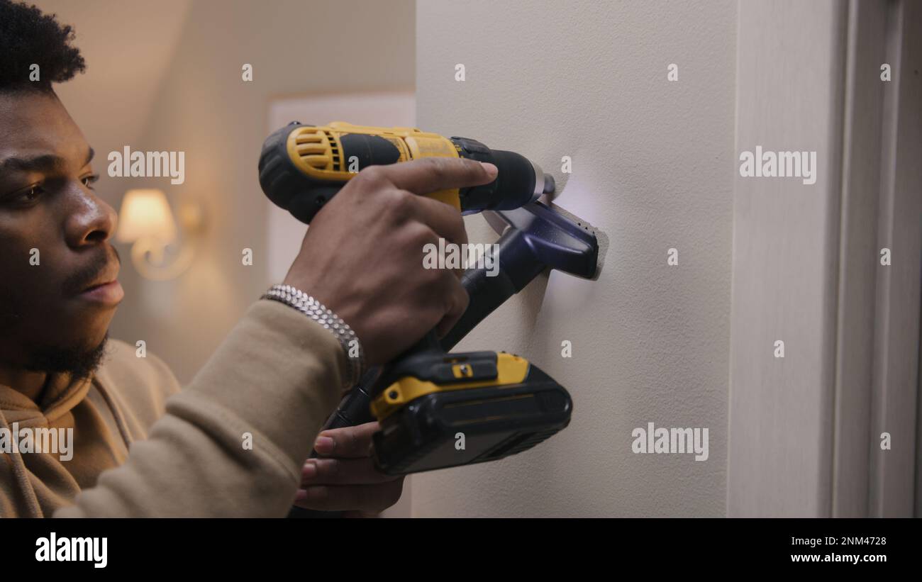 African American man in hoodie drills hole in concrete wall with hammer drill. Man does repairs ...