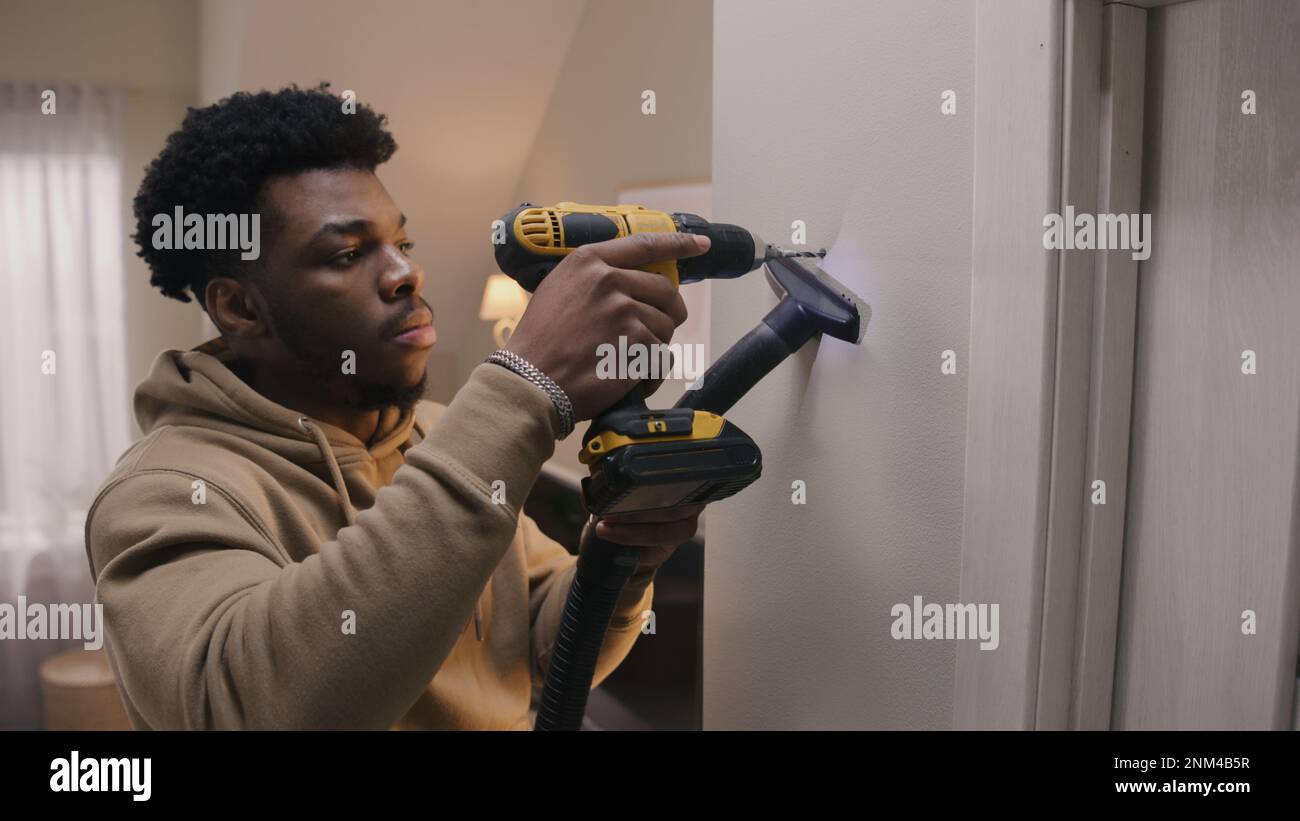 African American man in hoodie drills hole in concrete wall with hammer drill. Man does repairs ...