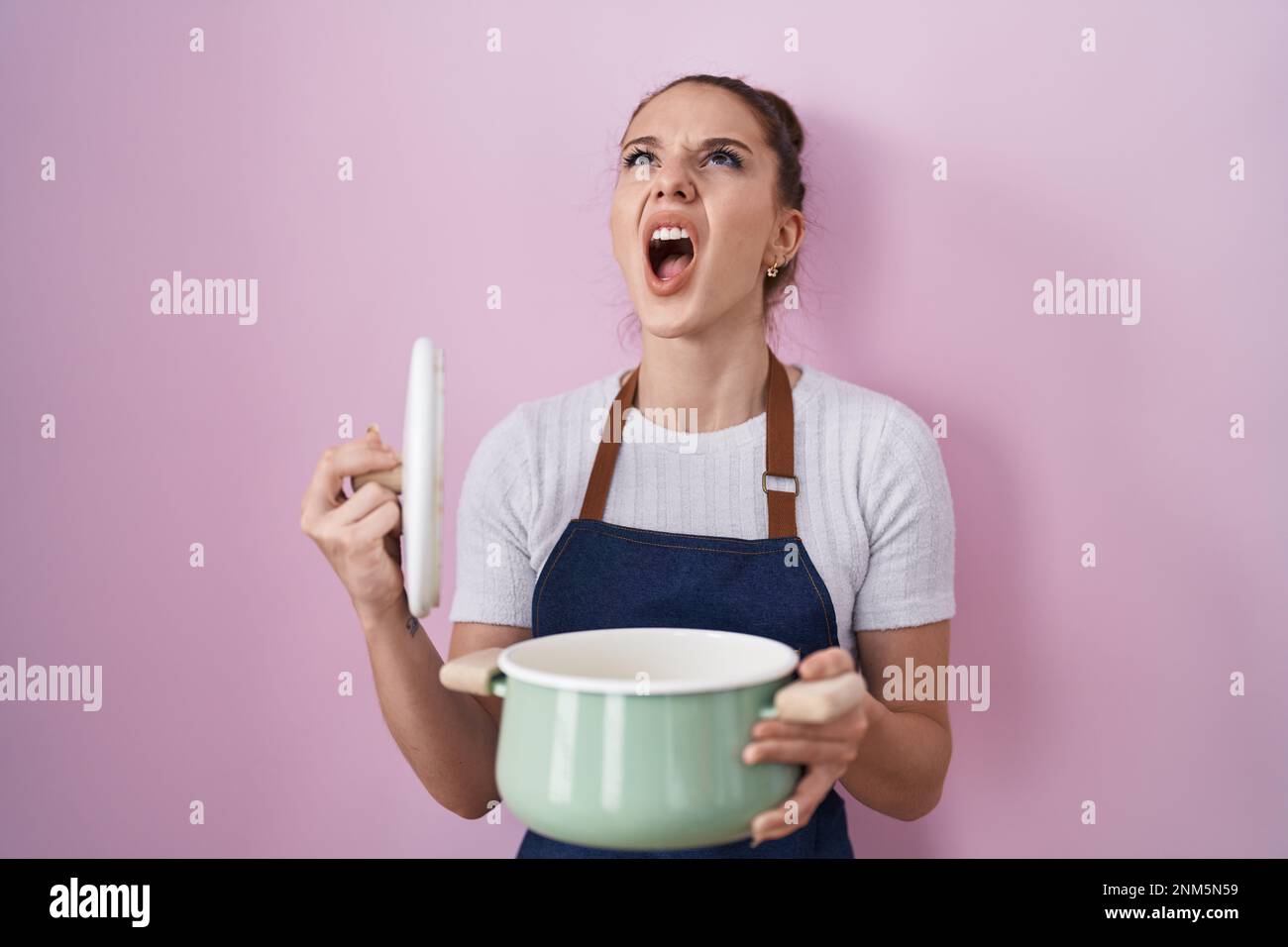 Young hispanic girl wearing apron holding cooking pot angry and mad ...