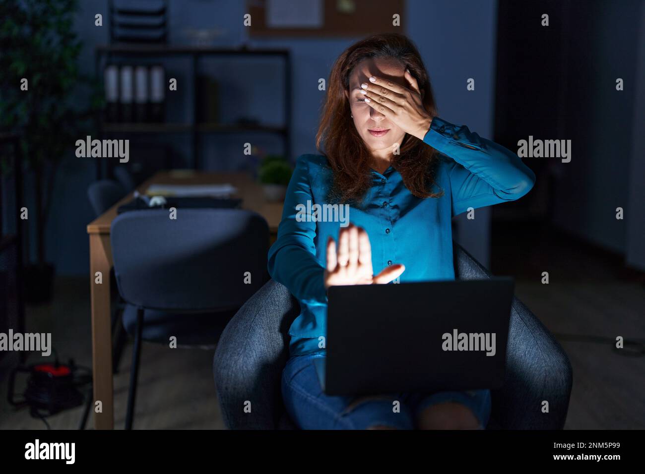 Brunette woman working at the office at night covering eyes with hands ...
