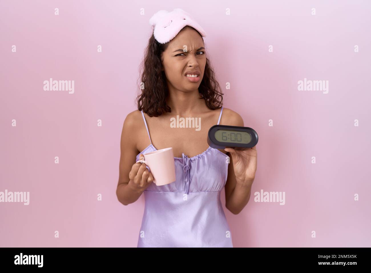 Young hispanic woman wearing nightgown holding alarm clock clueless and ...