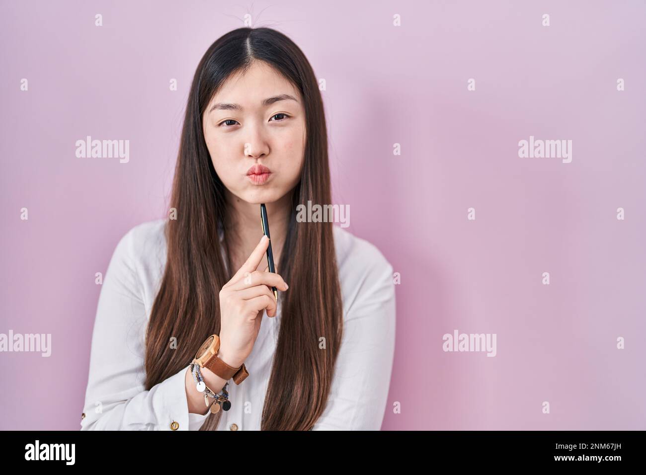 Chinese young woman holding pencil over pink background puffing cheeks with funny face. mouth ...
