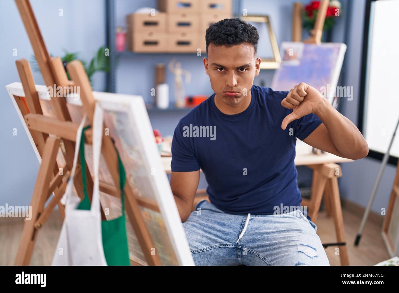 Young hispanic man painting sitting at art studio with angry face ...
