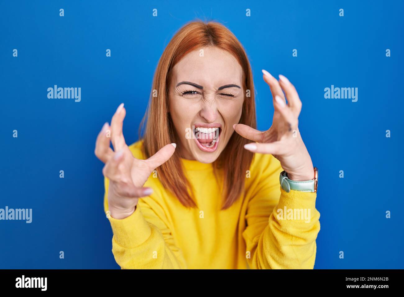 Young woman standing over blue background shouting frustrated with rage ...