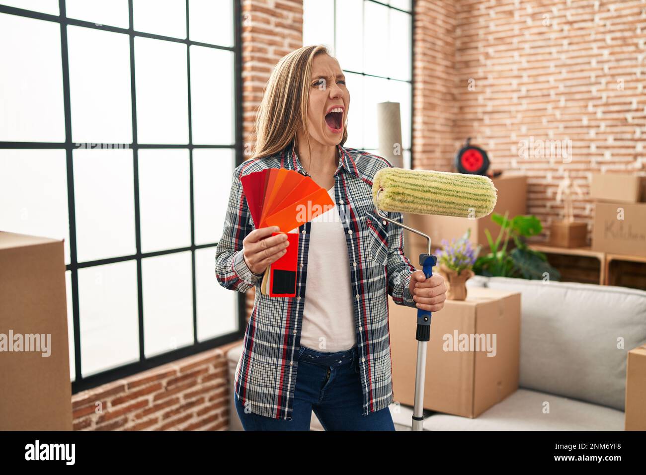 Young blonde woman holding roller painter and paint samples at new home ...