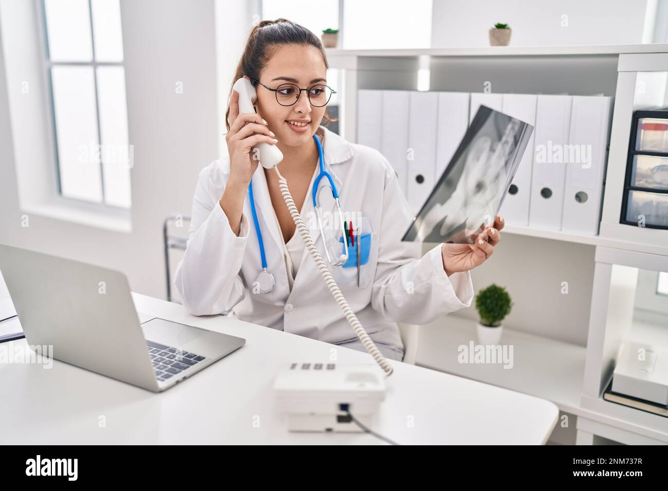 Young beautiful hispanic woman doctor talking on telephone holding xray ...