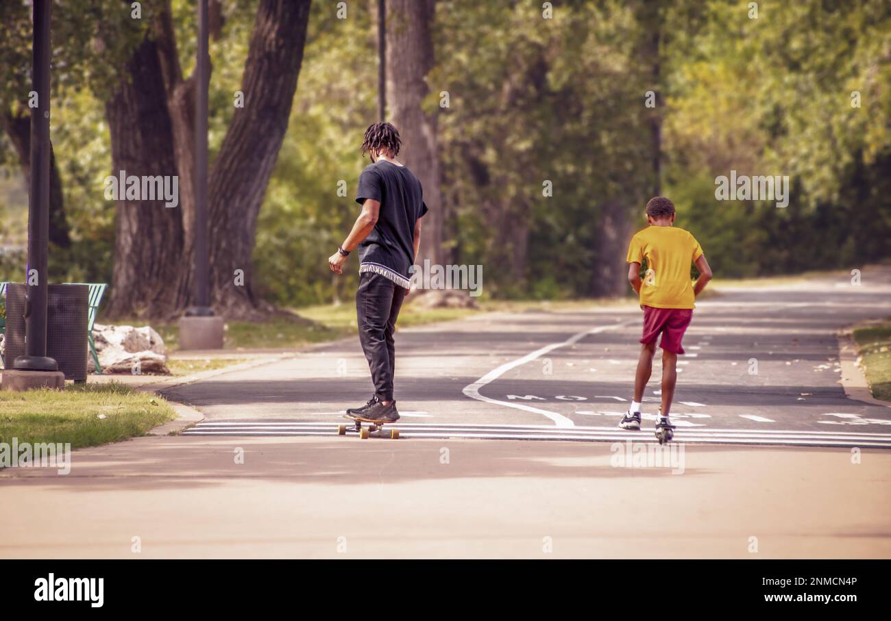 Two black boys one on scooter and one on skateboard on park trail with ...