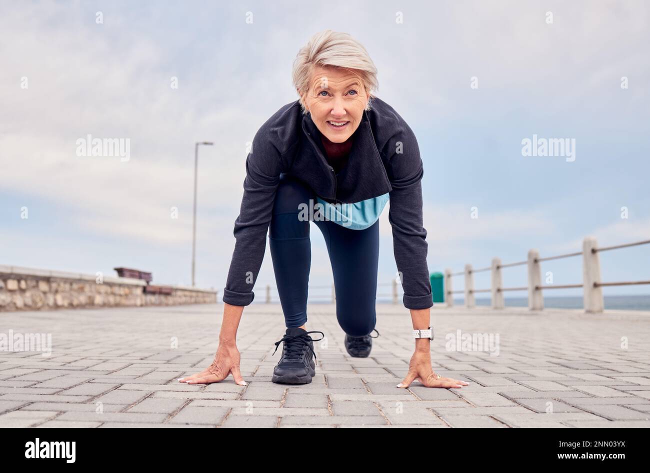 Senior woman, starting position and running outdoor at beach promenade ...
