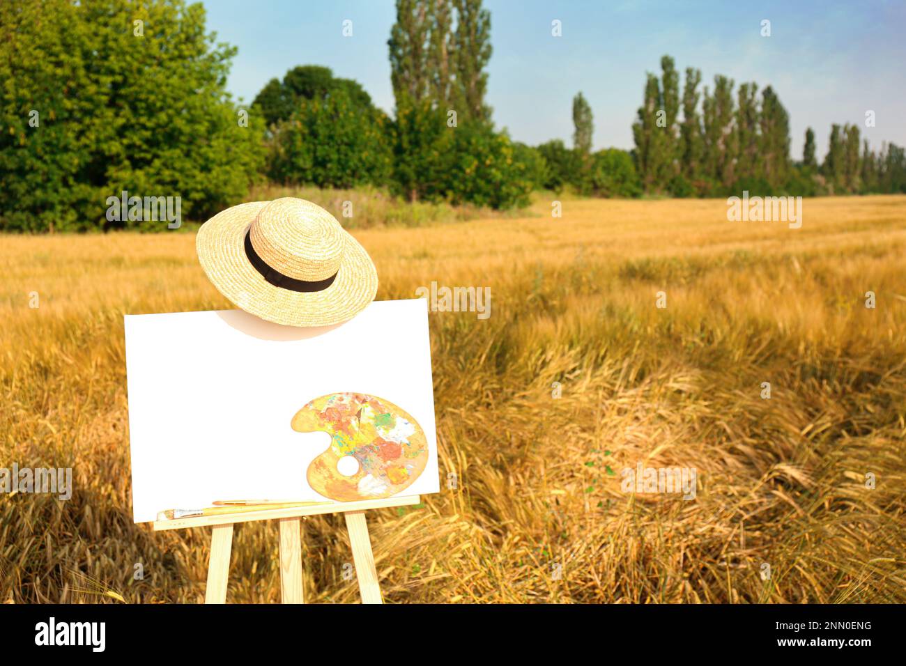Wooden easel with blank canvas, painting equipment and hat in field ...