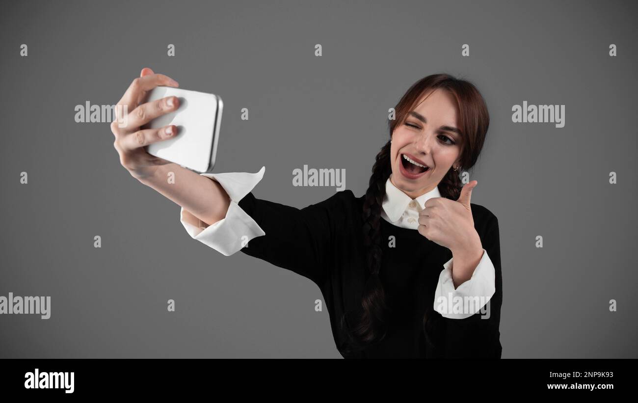 Smiling young girl student in black clothes with pigtails show thumb up sign, make selfie on ...