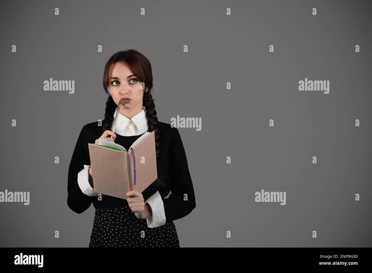 Pensive smart young girl student in gothic black clothes with pigtails with book, got idea and ...