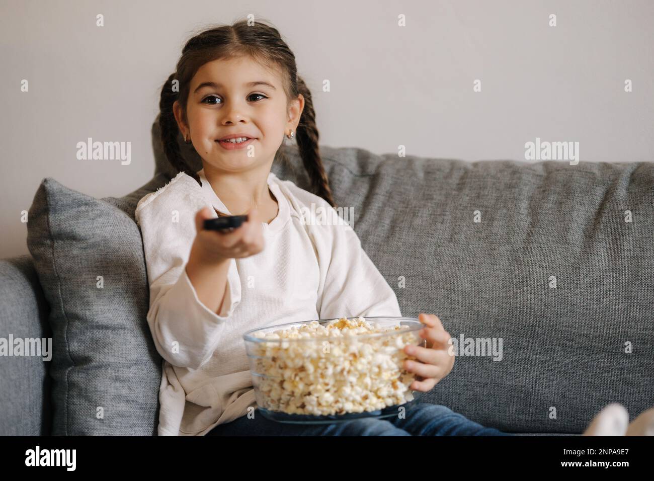 Smiled little girl using remote controller of TV and watching cartoons ...