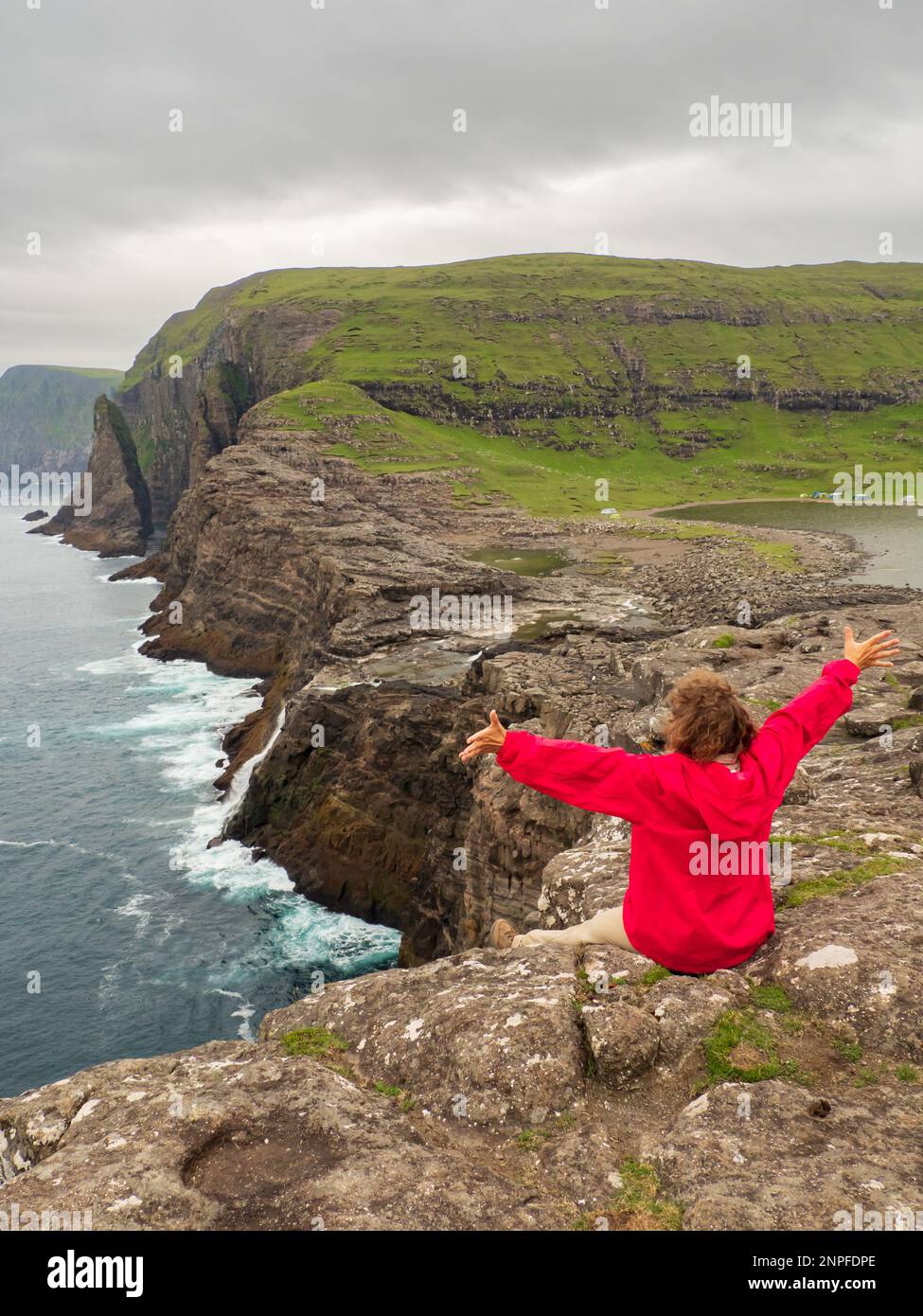 A tourist in a red jacket sits on a rocky shore above the Bøsdalafossur ...