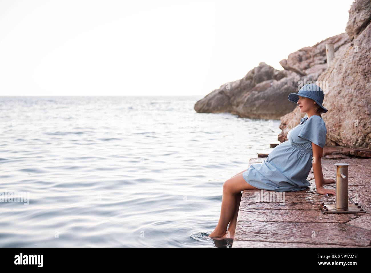 Pregnant woman traveler in denim dress, blue straw hat sits by the sea ...