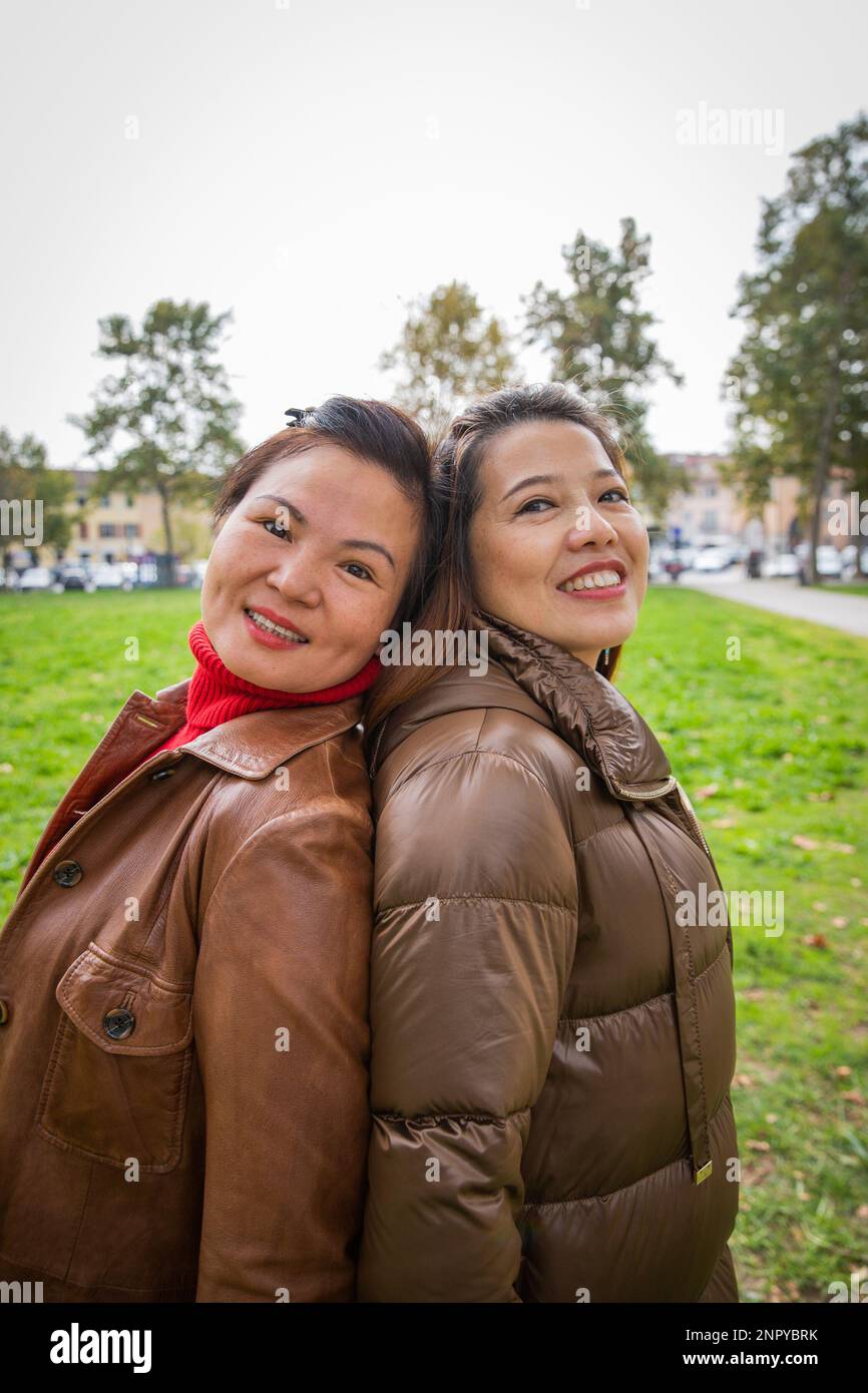 Two smiling friendly Asian women pose back to back in a public park ...