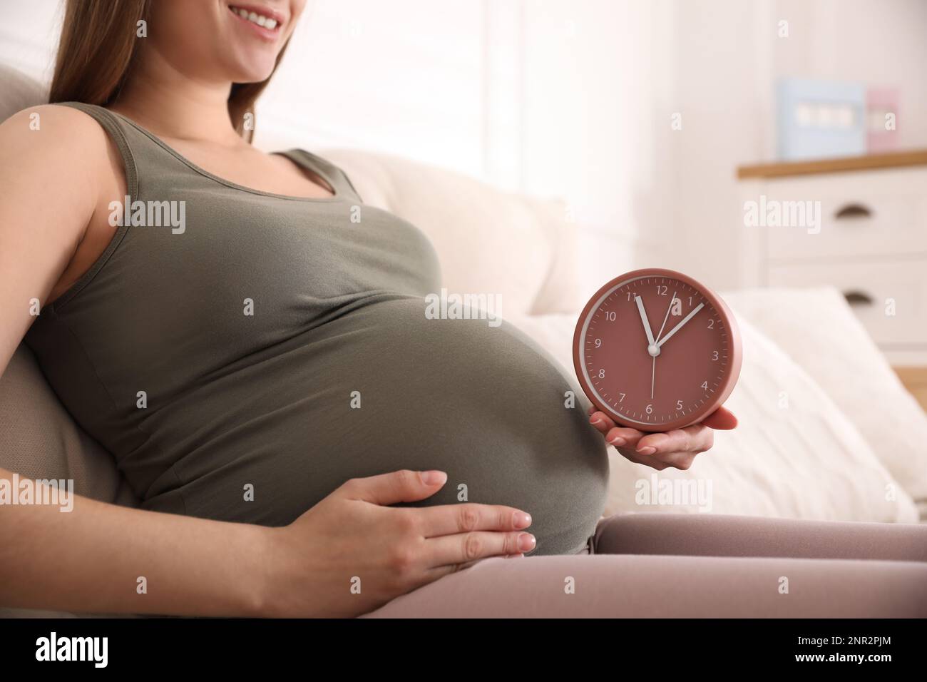 Young pregnant woman holding clock near her belly at home, closeup ...