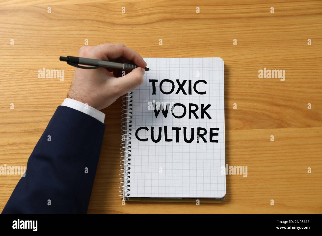 Man writing text Toxic Work Culture in notebook at wooden table, top ...