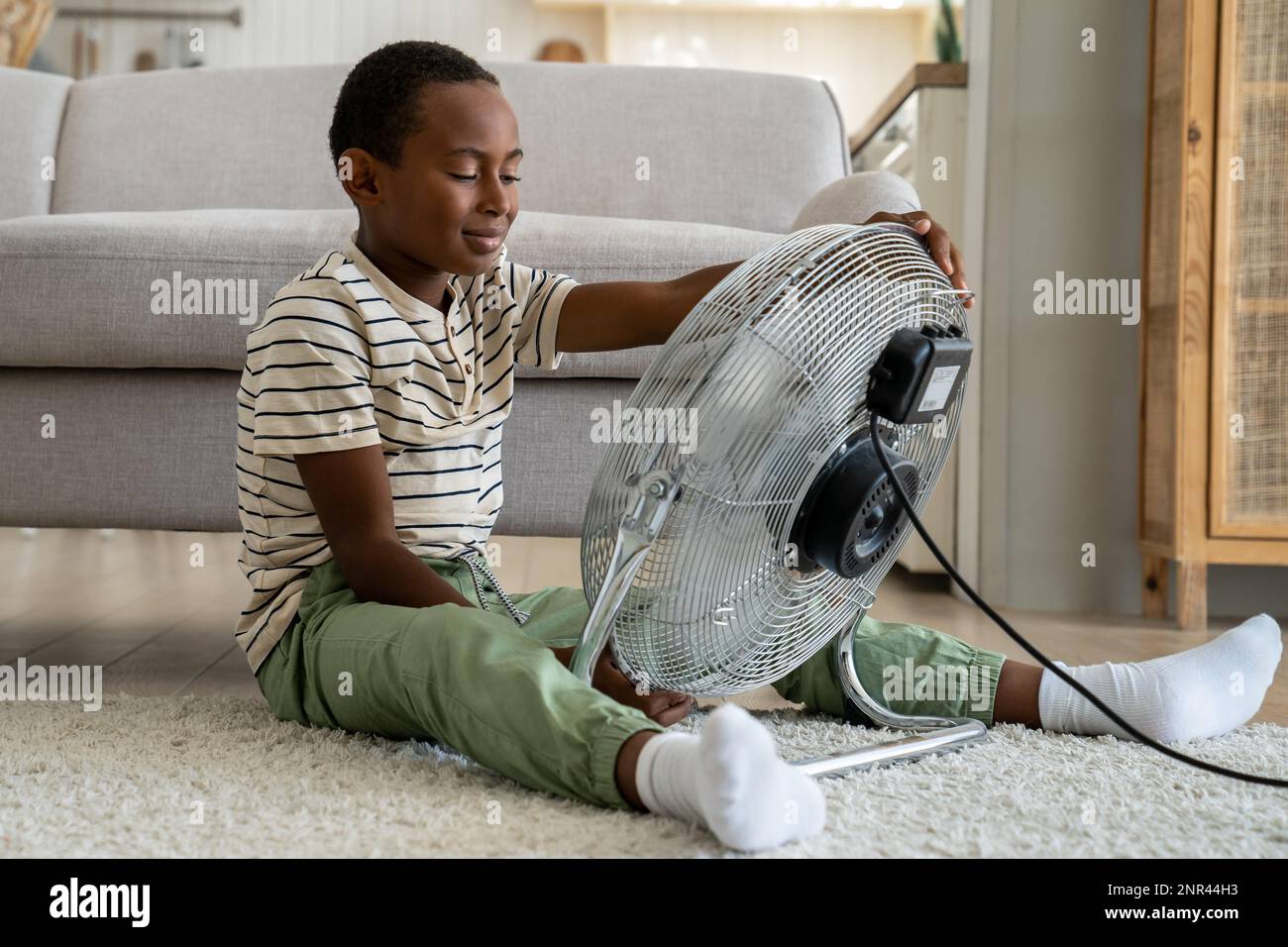 Calm African boy refreshing with electric fan at home alone. Small boy ...