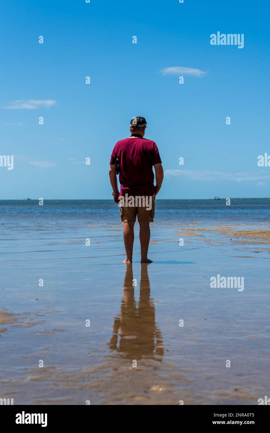 Back view of man standing on the wet beach at low tide, looking out to ...