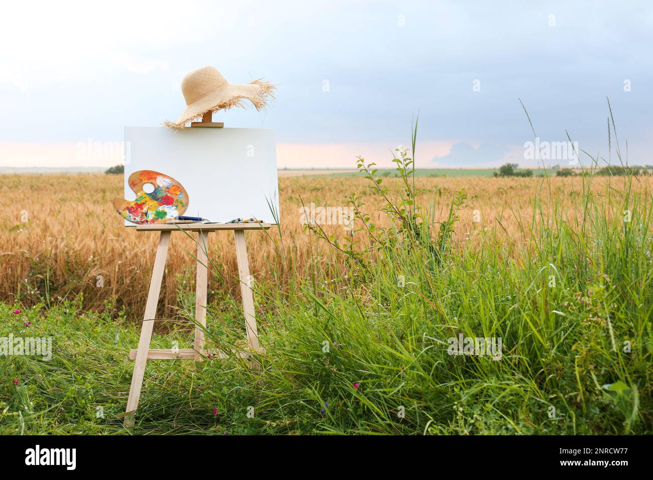 Wooden easel with blank canvas, painting equipment and hat in field ...