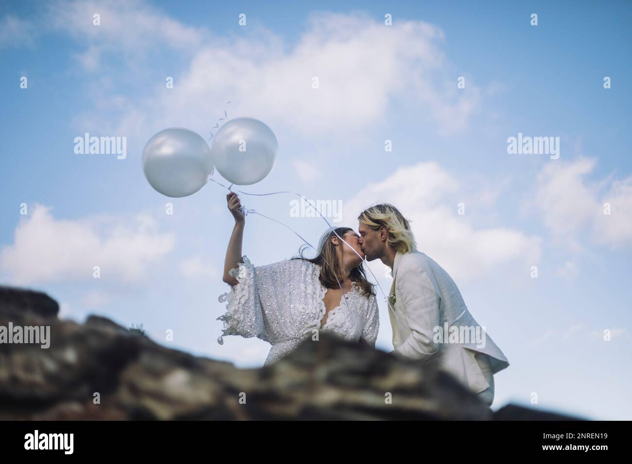 Romantic bride holding white helium balloons kissing groom against sky ...