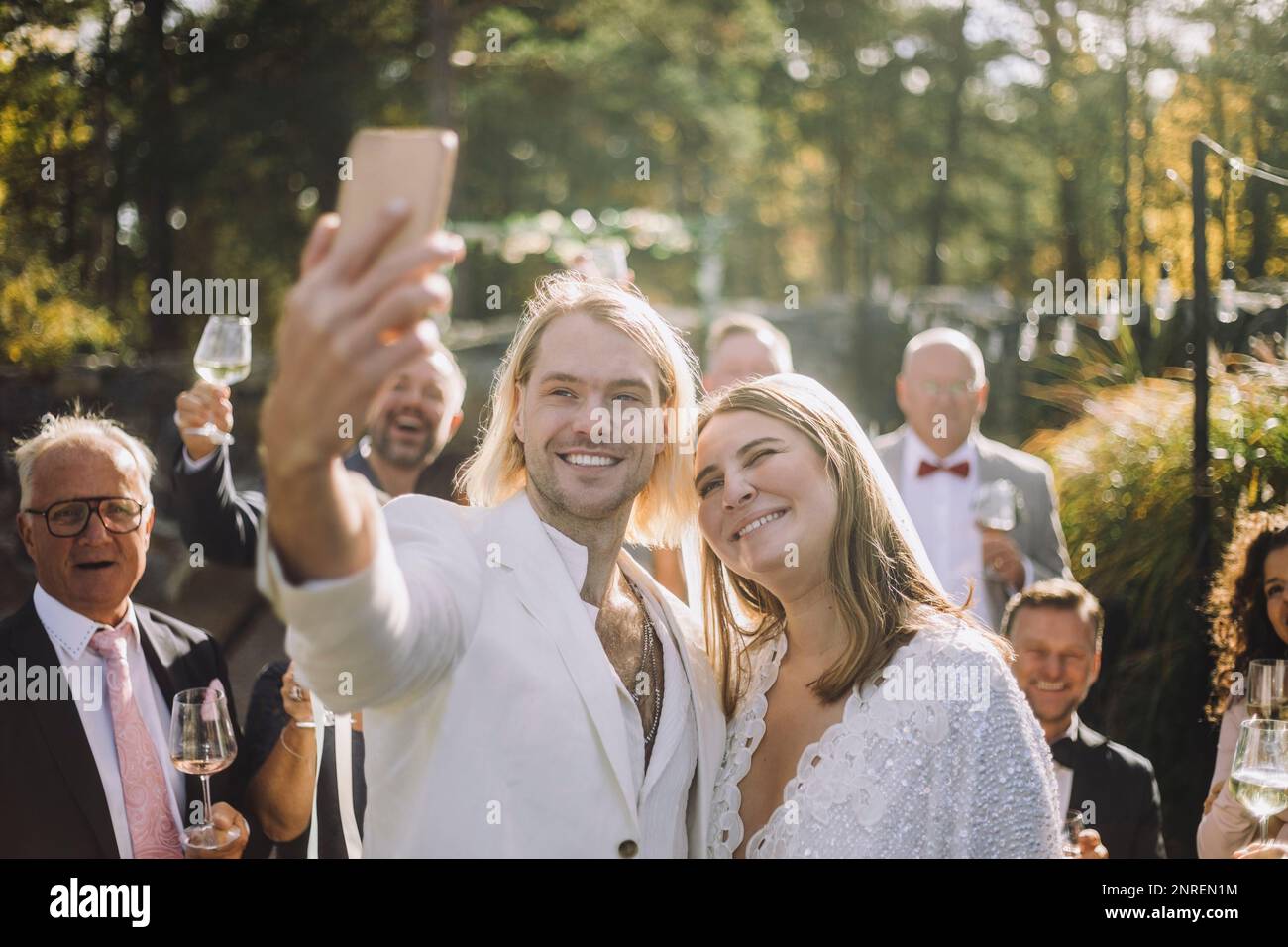 Happy groom taking selfie with bride and guests through smart phone at ...