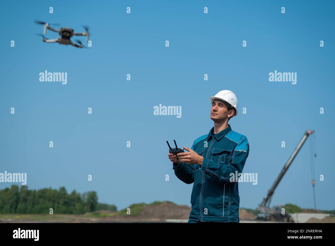 A man in a helmet and overalls controls a drone at a construction site ...