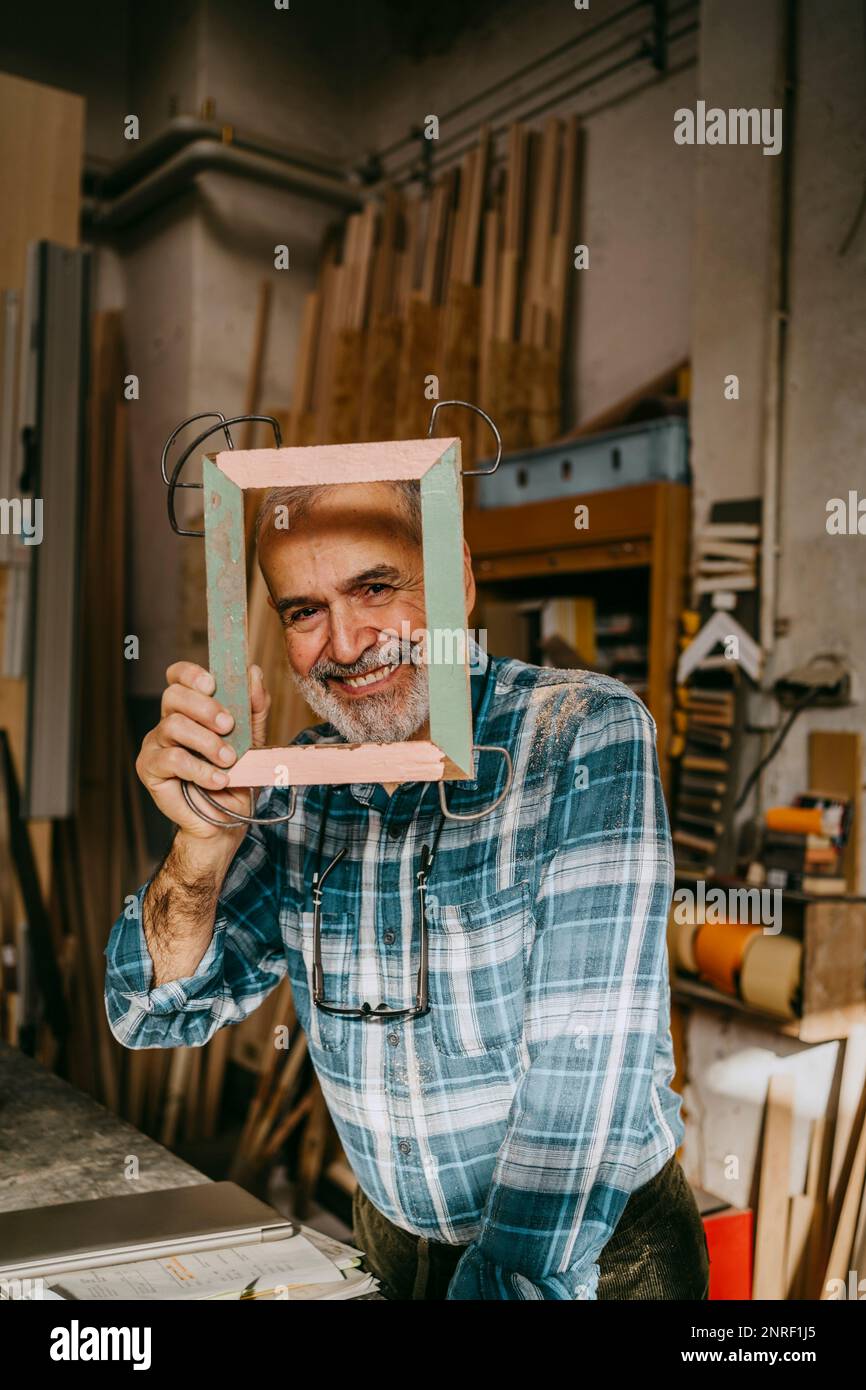 Portrait of happy craftsman looking through picture frame at workshop ...