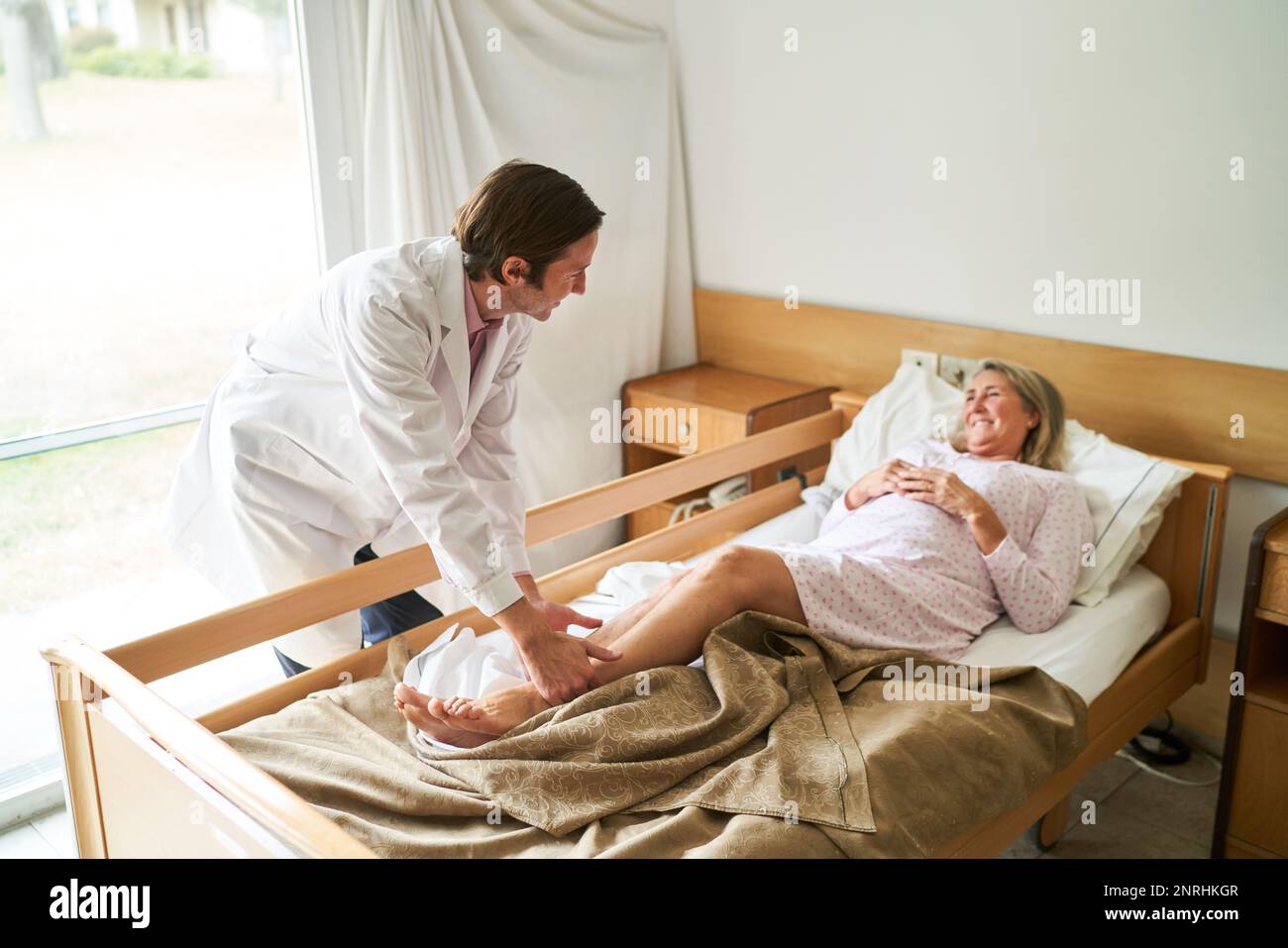 Smiling male doctor lifting legs of elderly female patient lying in bed ...