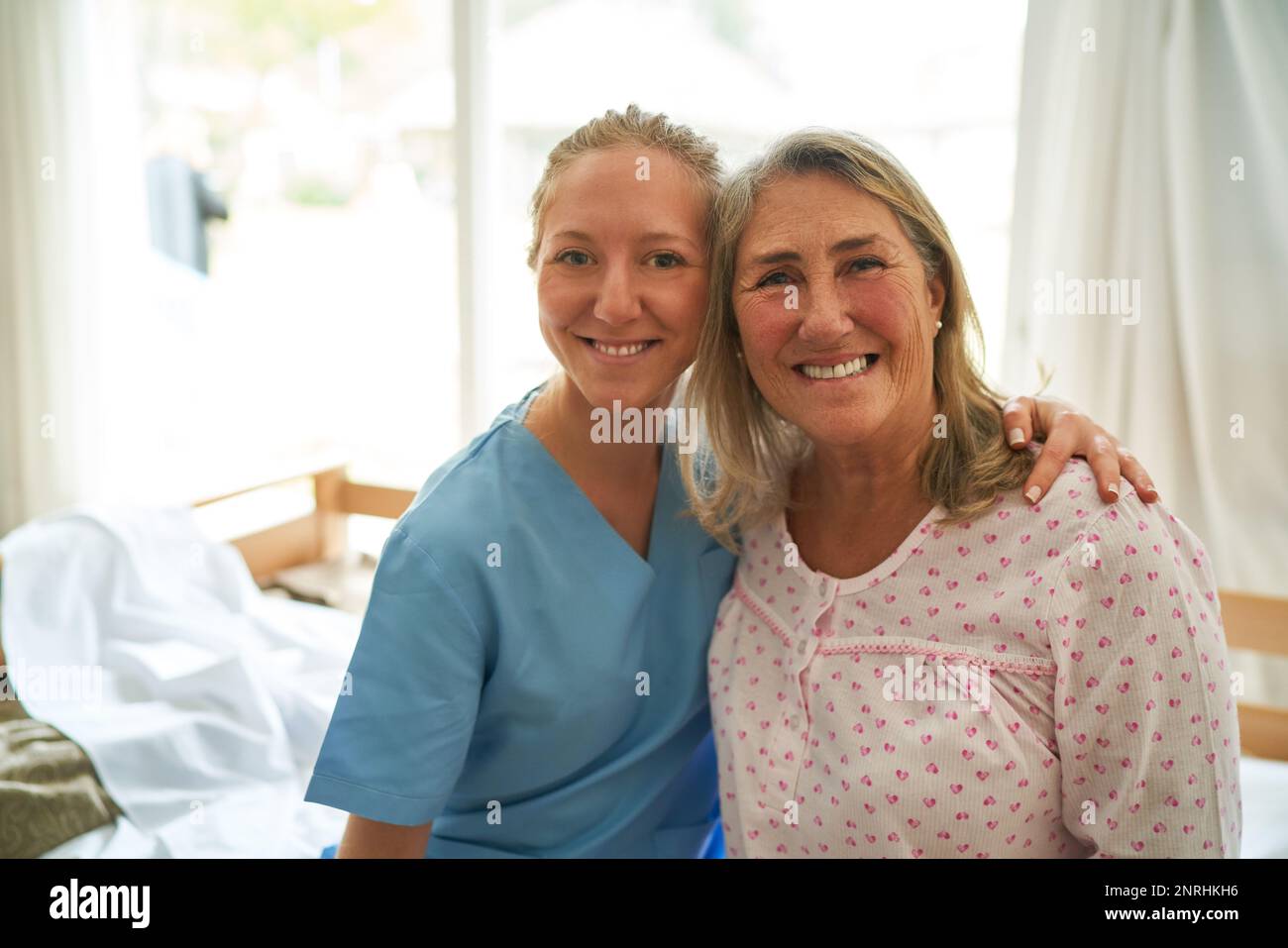 Portrait of smiling caring nurse embracing elderly woman sitting on bed ...