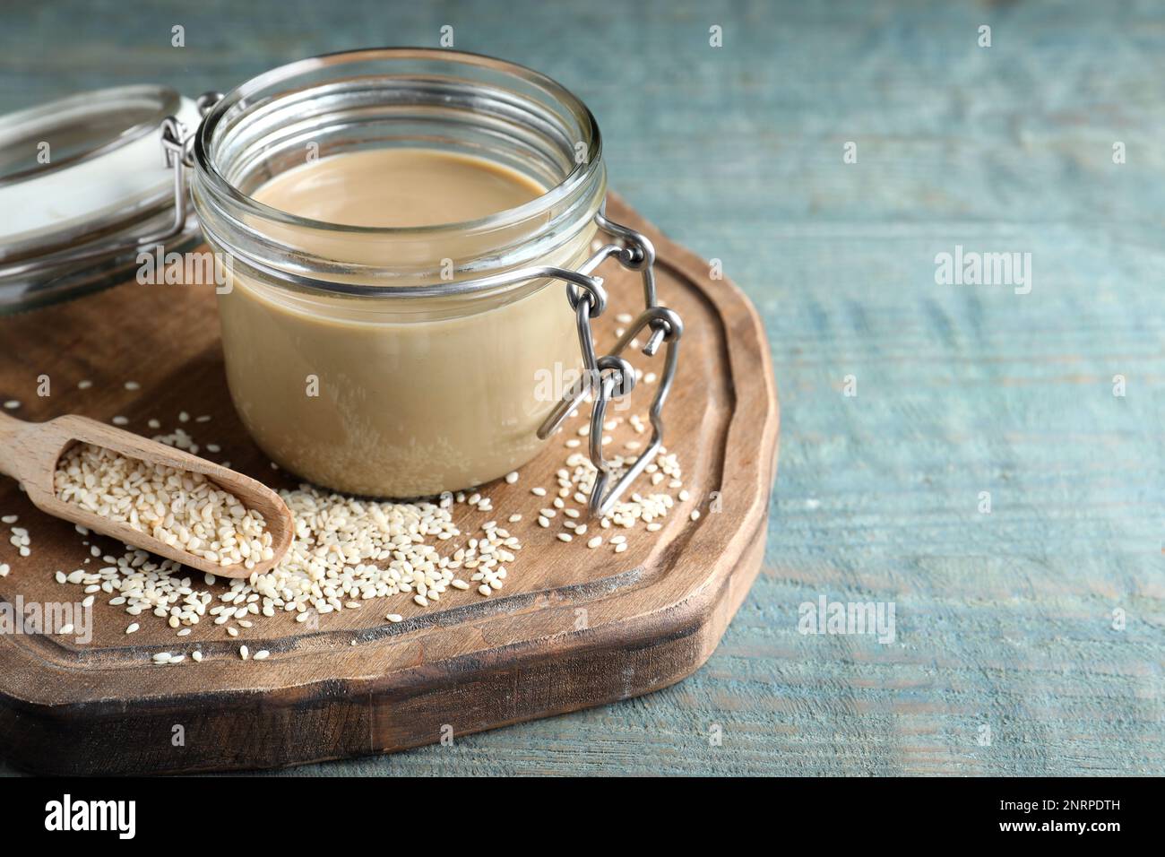 Jar of tasty sesame paste, seeds and scoop on light blue wooden table ...
