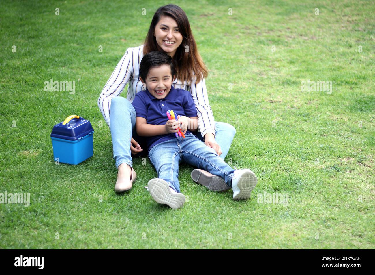 Latino mom and 6 year old boy sitting on the grass with lunch box and ...
