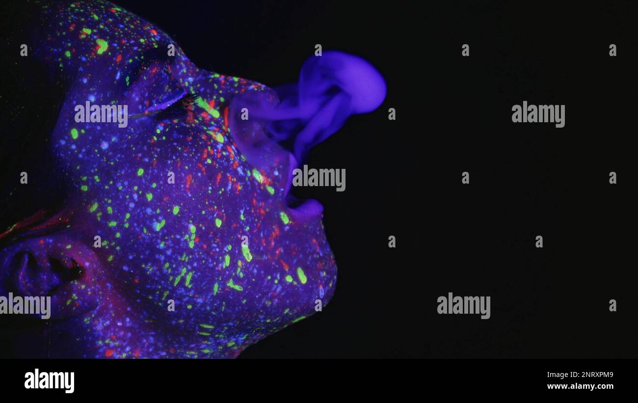 A beautiful young girl with makeup from ultraviolet paints smokes ...