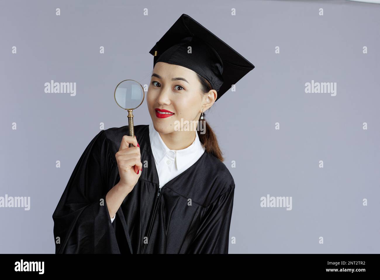 modern female asian graduate student with magnifying glass against grey ...
