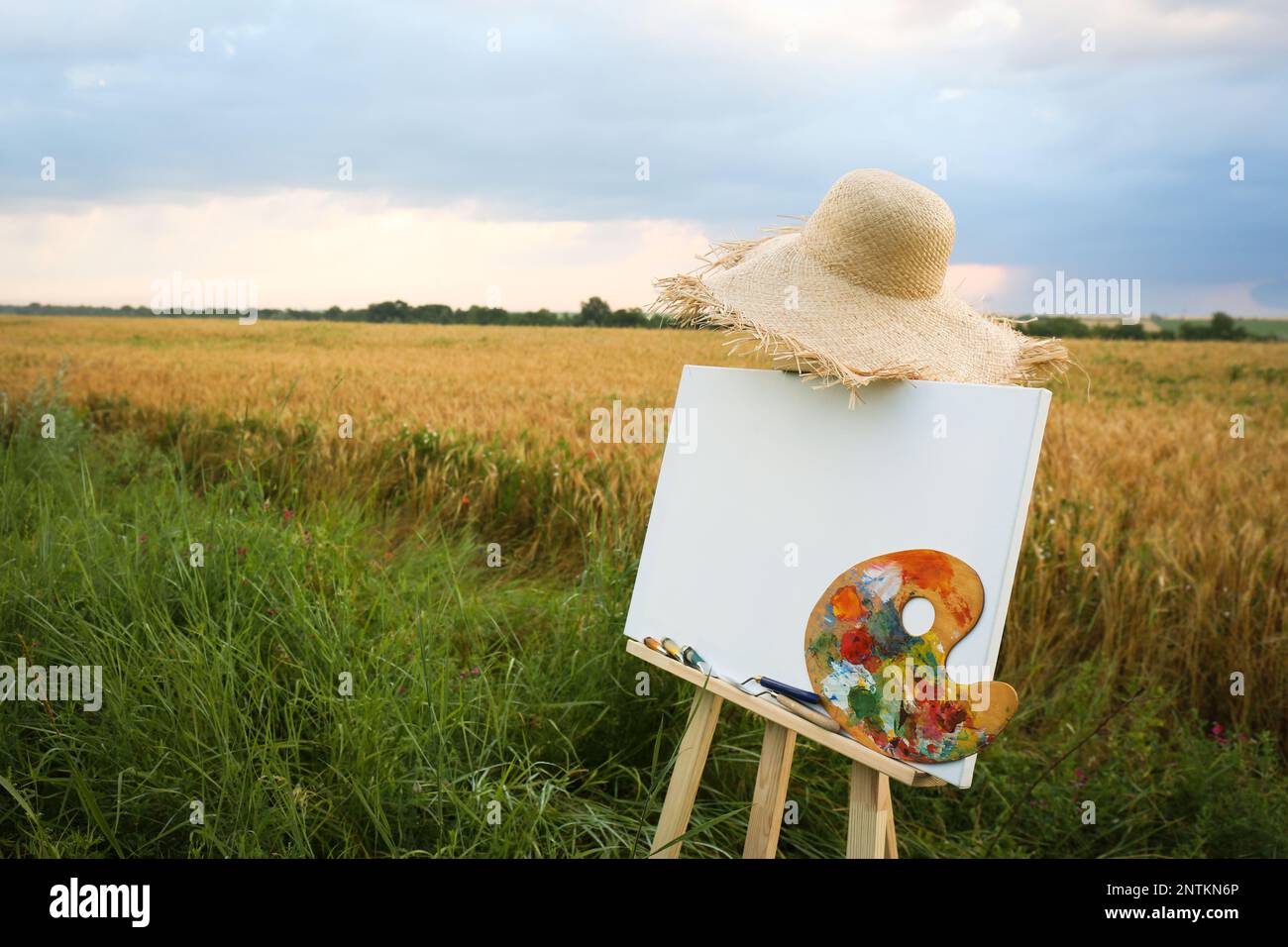 Wooden easel with blank canvas, painting equipment and hat in field ...