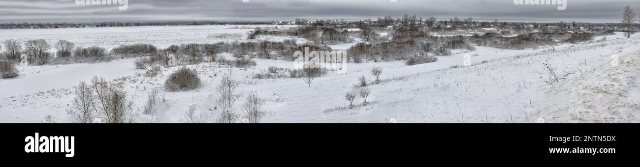 Winter panorama with a frozen river with bushes and a small village ...