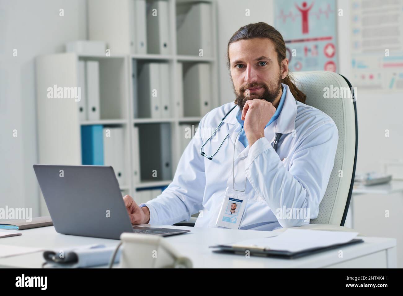 Young successful male clinician in lab coat sitting by workplace in ...