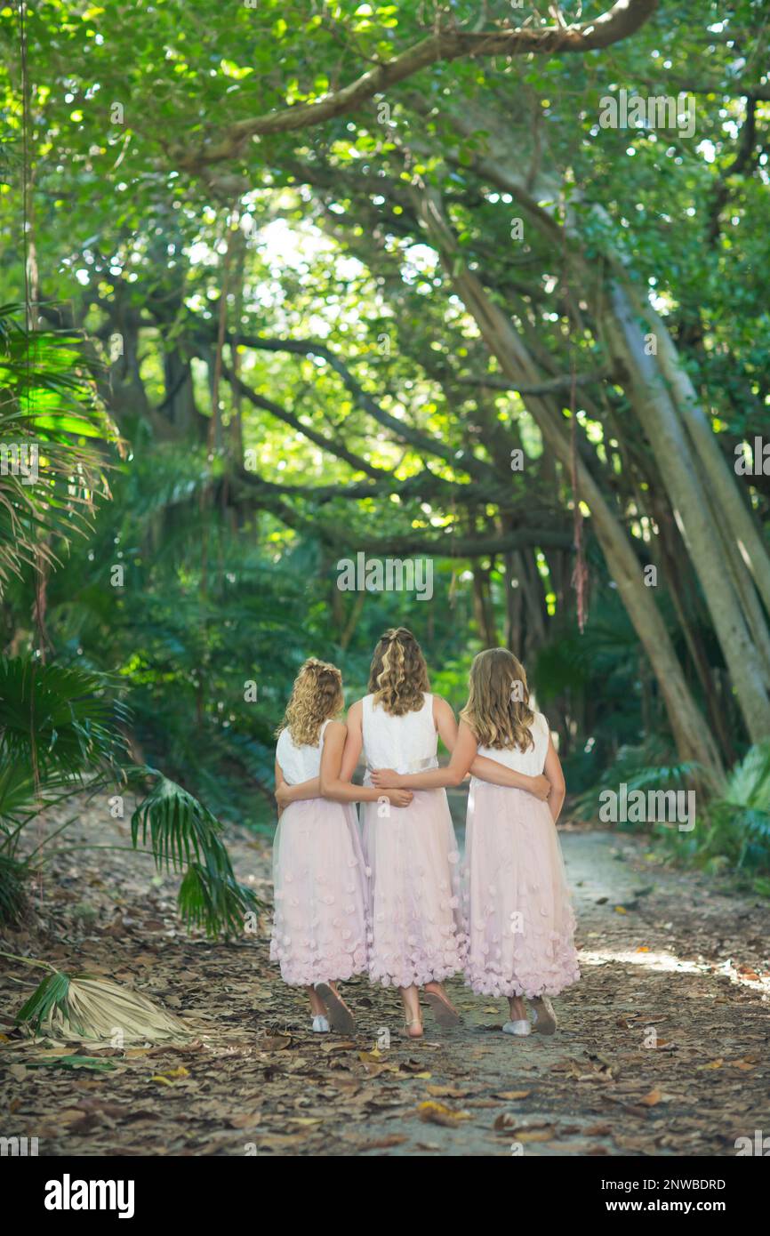 three sisters in pale pink dresses walking through tunnel of trees arm ...