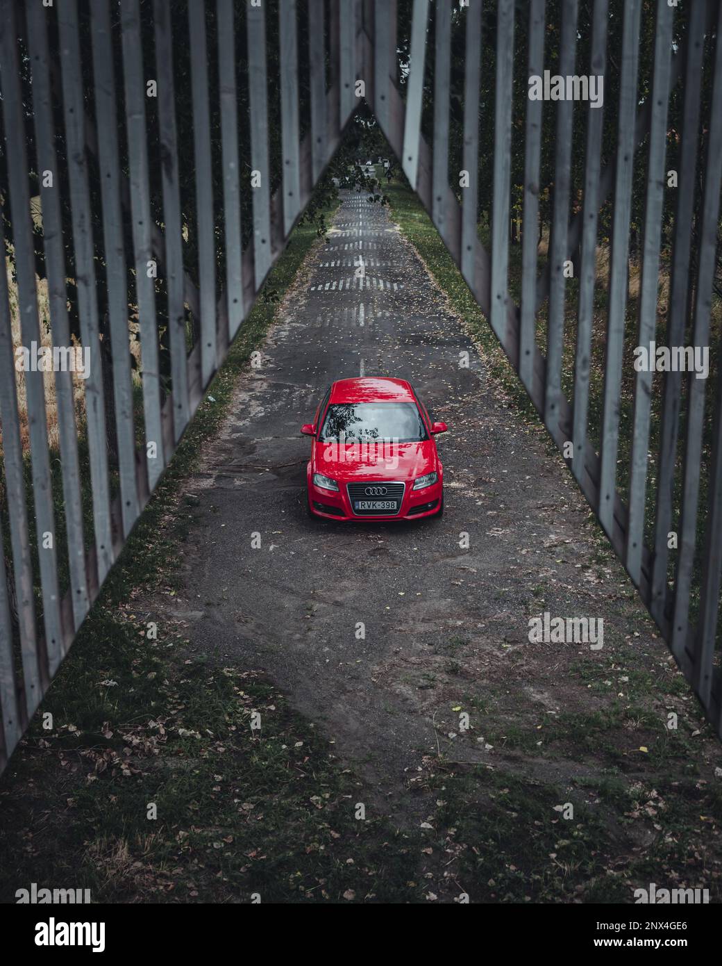 A red sports car is parked on a dirt path next to a tall wooden fence ...