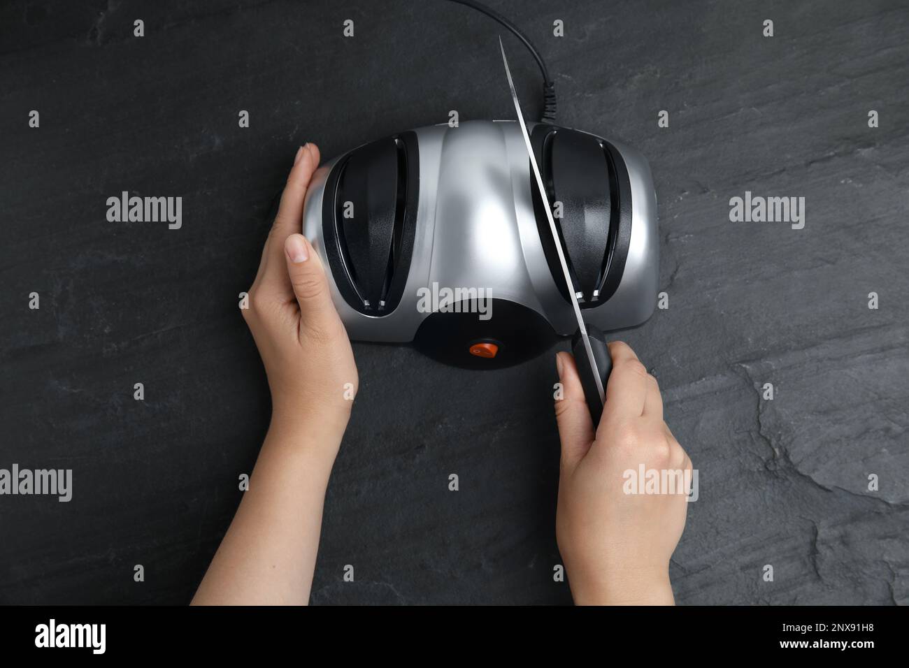 Woman sharpening knife with electrical tool at black table, top view ...