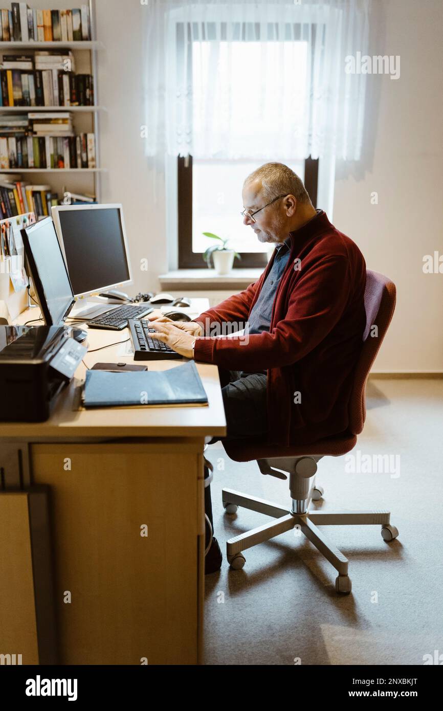 Full length of senior man typing while using computer at desk in home ...