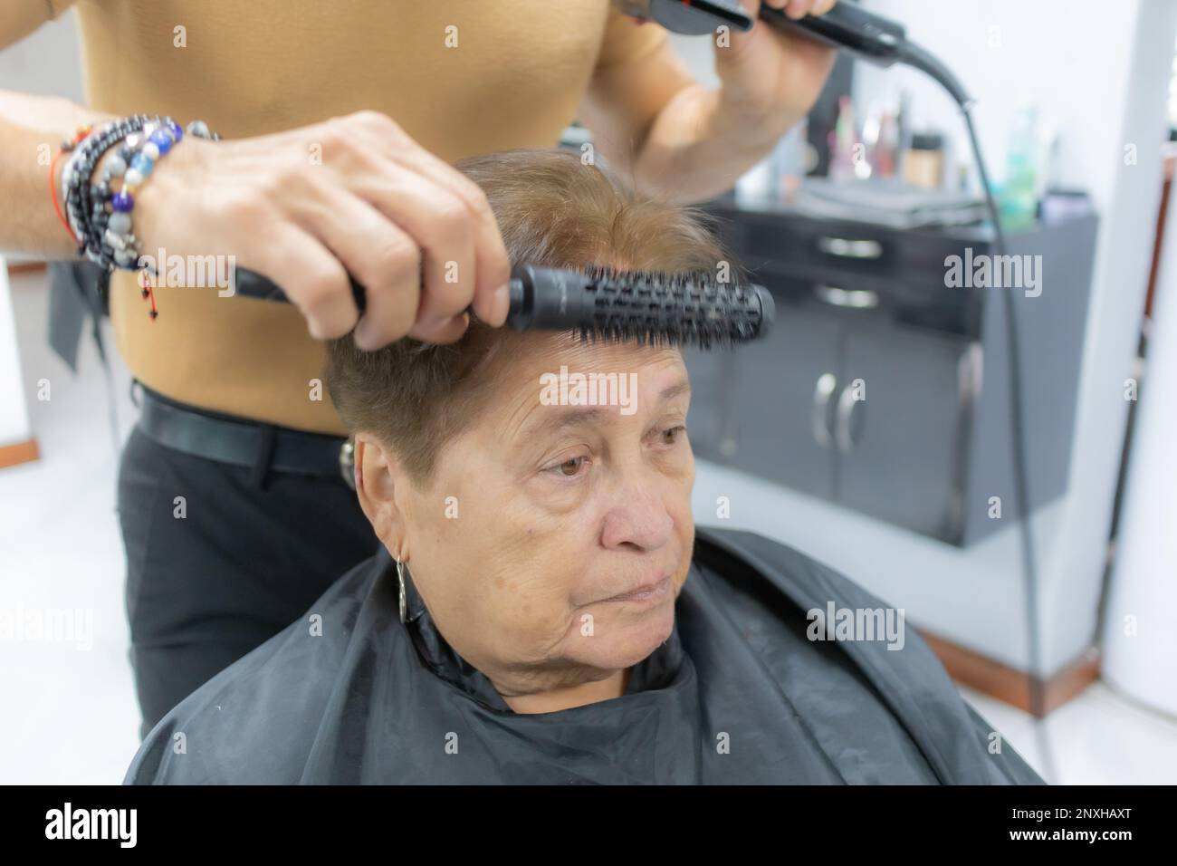 Senior woman in beauty salon, stylist styling forelock with hair dryer ...