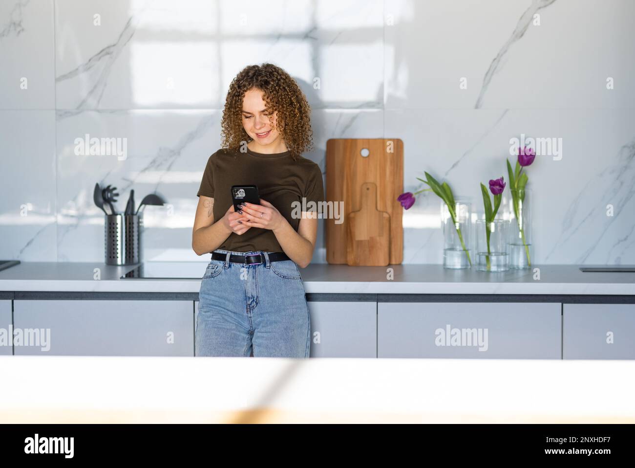 Beautiful young woman standing by kitchen counter reading text message ...