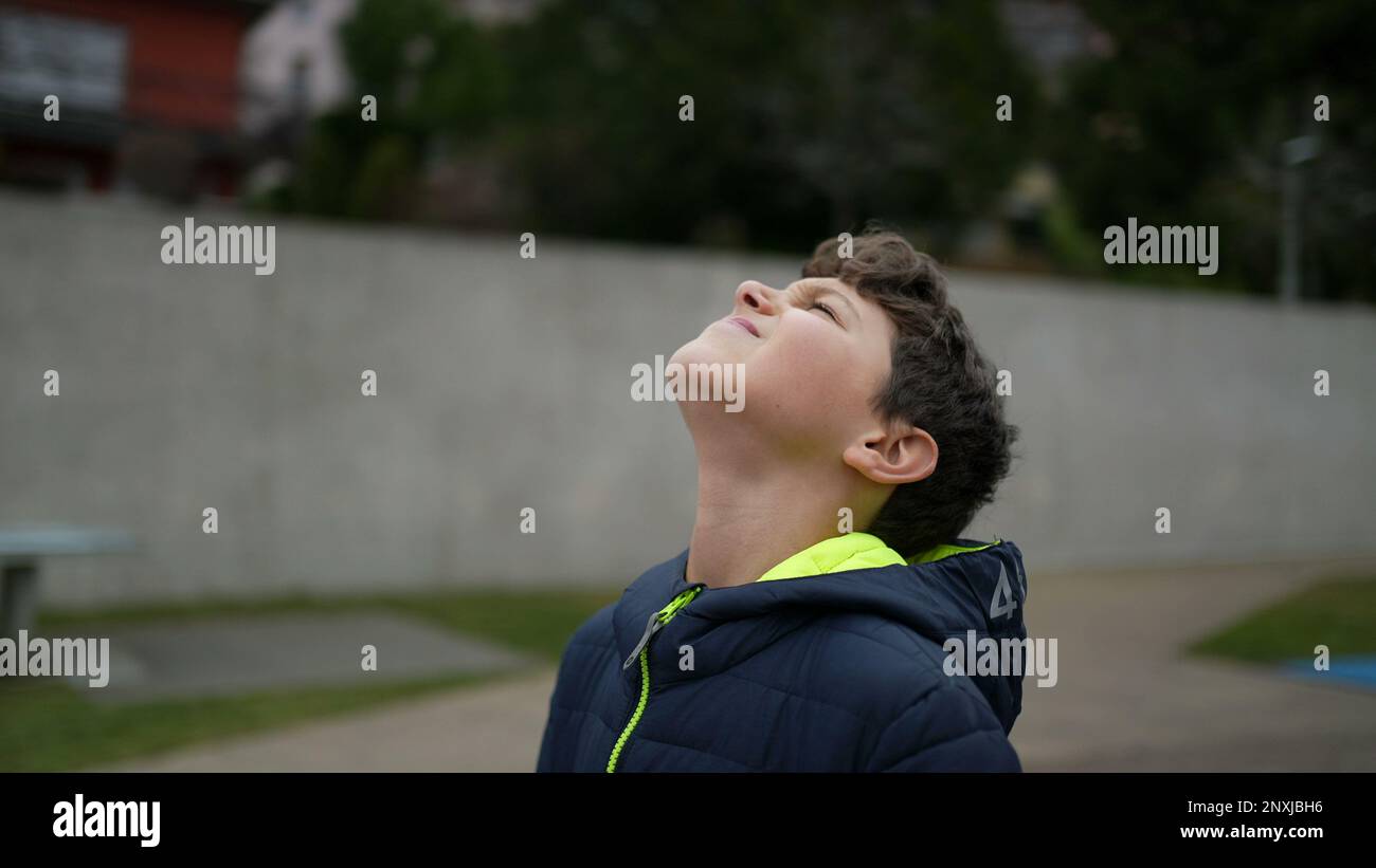 One contemplative young boy looks up at sky. Kid standing outside at ...